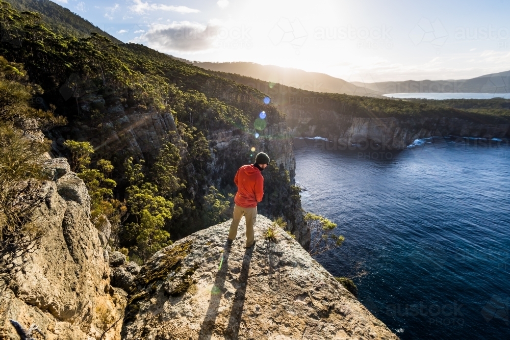 Image of A man stands on a cliff overlooking the ocean - Austockphoto