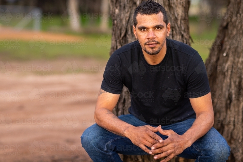 Image of aboriginal man casually squatting down in front of tree trunk ...