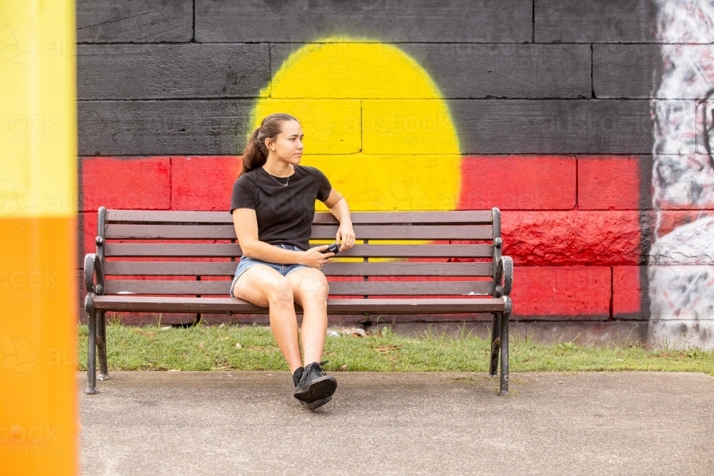 Image of Aboriginal woman sitting on a park bench in front of a mural ...