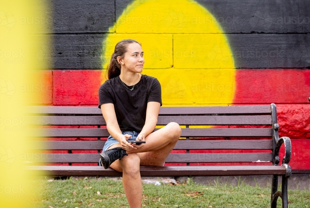 Image of Aboriginal woman sitting on a park bench in front of a mural ...