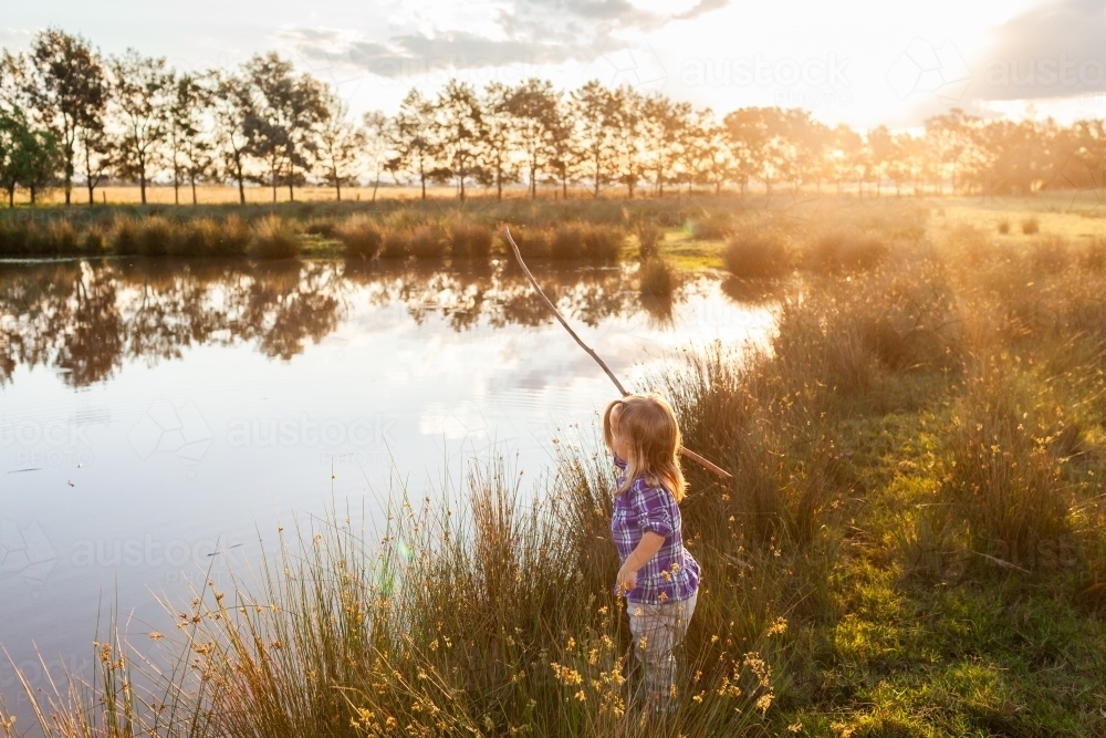 Image of Aussie child in golden light playing by farm dam with stick ...