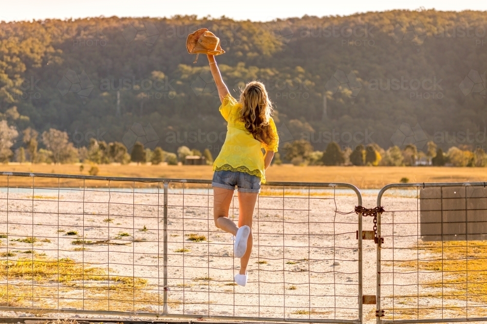 Image of Aussie woman standing on a rural farm gate with one arm raised ...