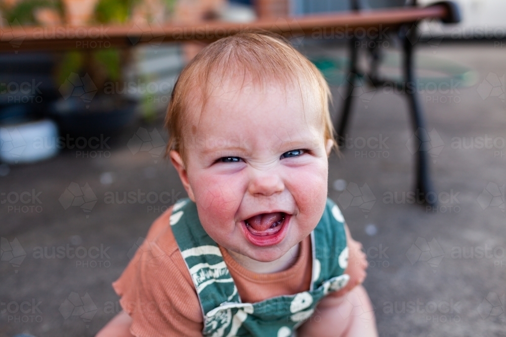 Image of Baby laughing and pulling face outside playing in overalls ...