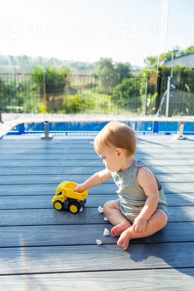 Image of baby sitting on back deck beside glass pool fence playing with ...