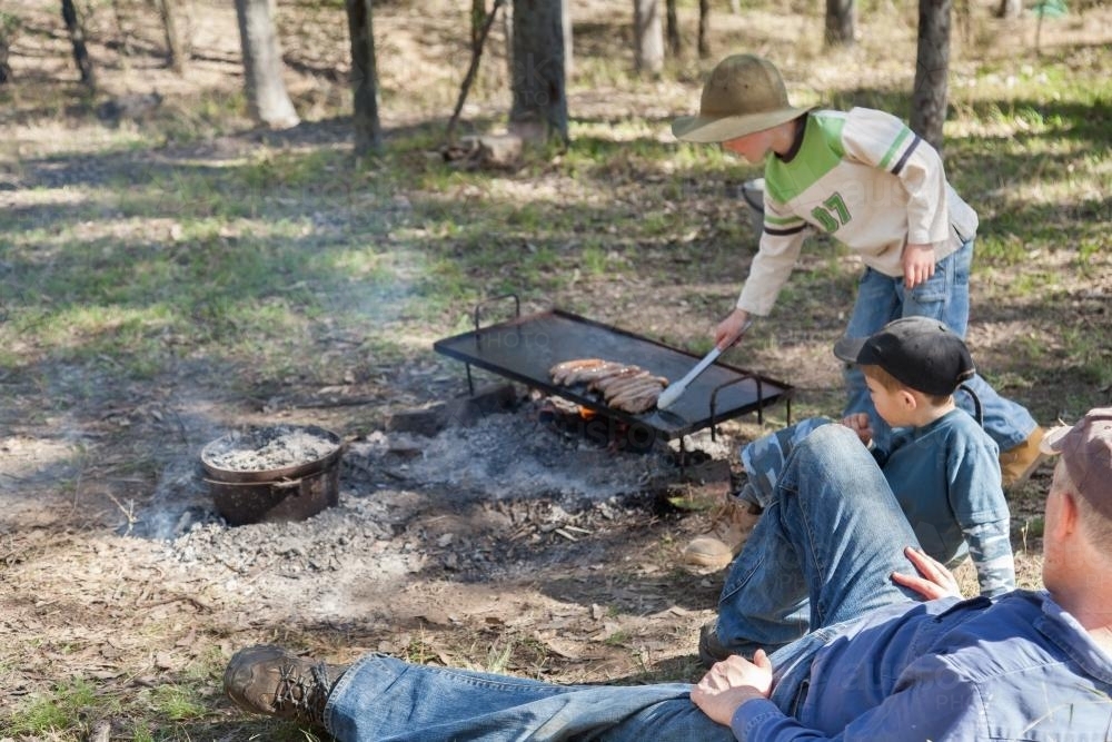 Image of Boys cooking and relaxing around the campfire - Austockphoto