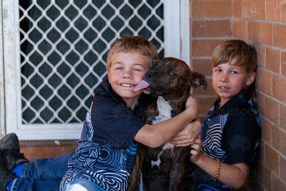 Image of Happy Aussie kid sitting at doorstep of home with dog licking ...