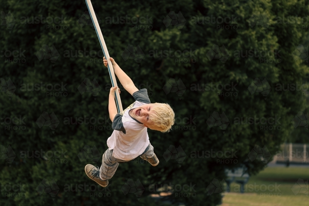 Image of Happy boy playing on swing at playground - Austockphoto