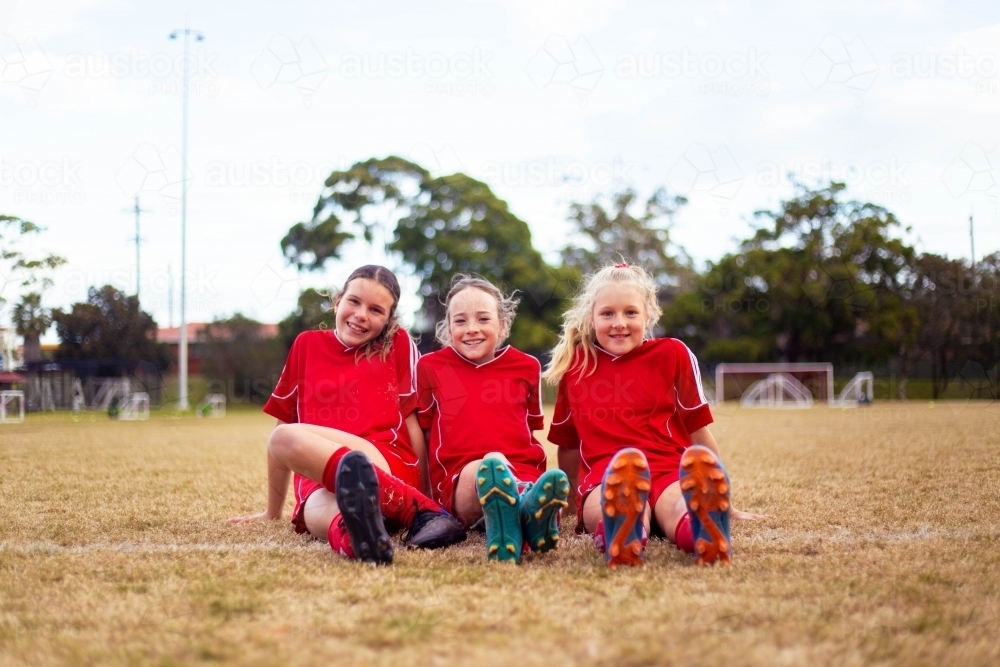 Image of Happy girls wearing red soccer uniforms sitting on the grass ...