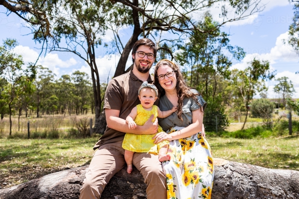 Image of Happy smiling young family of three sitting on log in farm ...