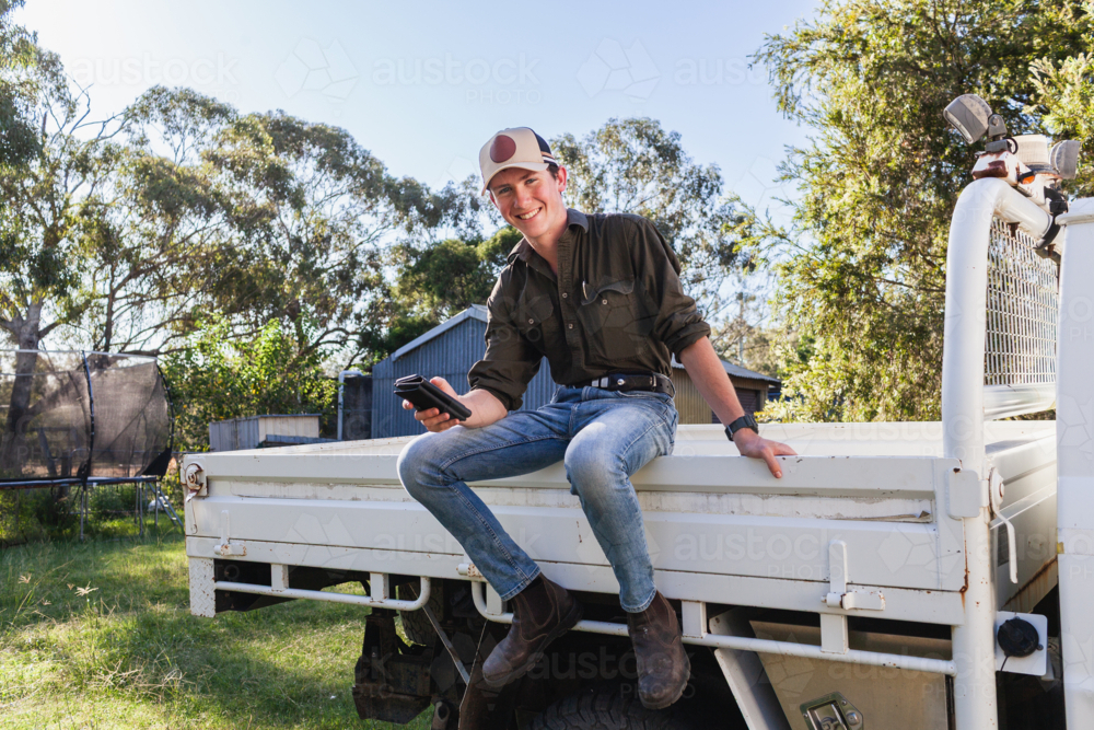 Image of happy teenaged young man sitting on ute tray using mobile ...