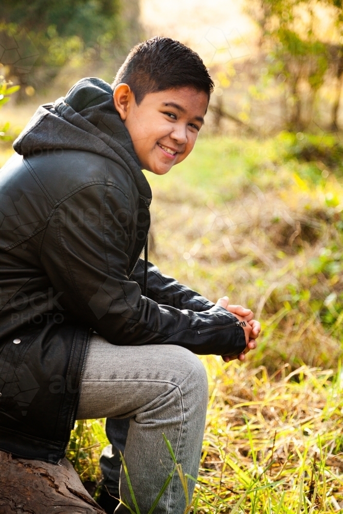 Image of Happy tween boy sitting on log outside smiling - Austockphoto