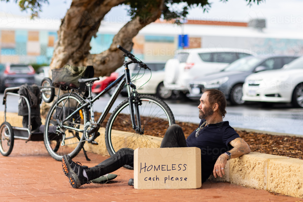 Image of homeless man sitting on footpath near a carpark with his bike ...