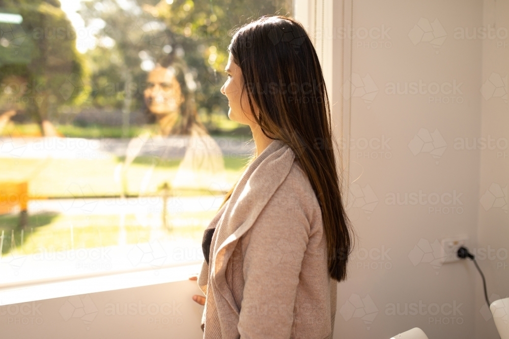 Image of Indian woman standing by the window looking outside on a sunny ...