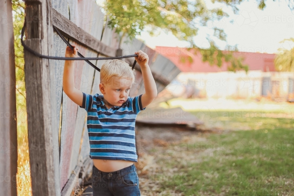 Image of Little boy on farm pulling on rubber attached to tin fence ...