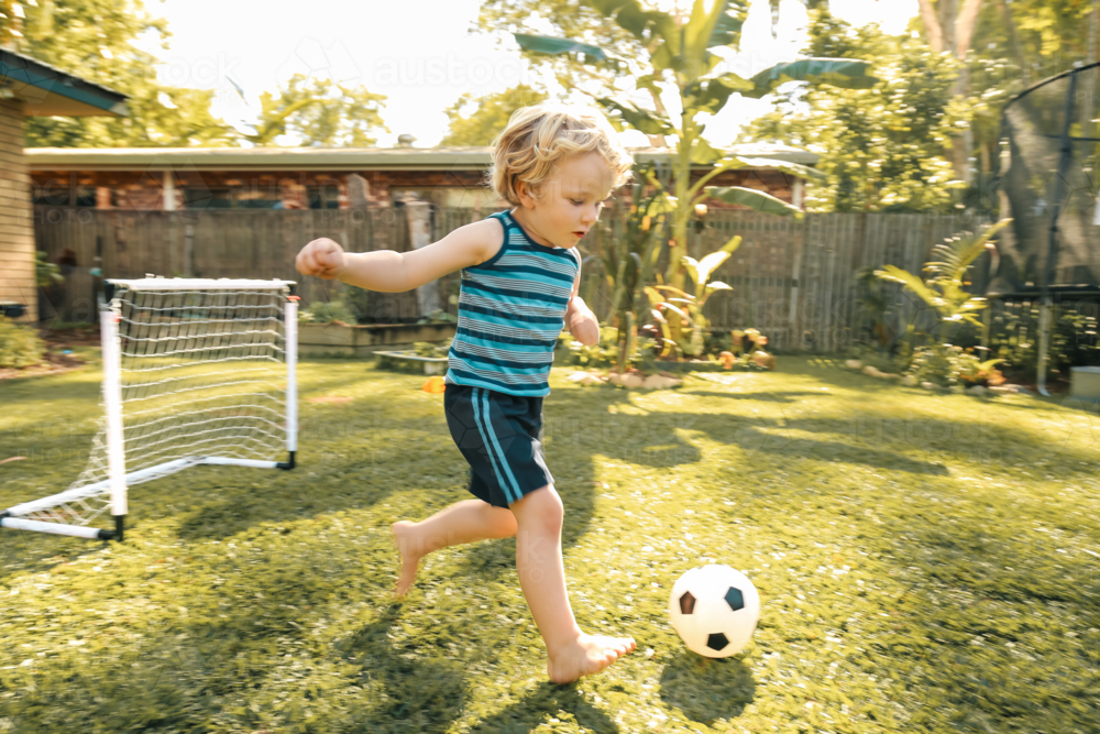 Image of Little boy playing soccer in sunlit backyard - Austockphoto