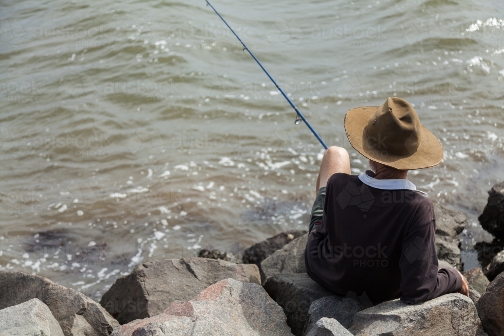 Image of Man fishing off rocks beside the sea - Austockphoto