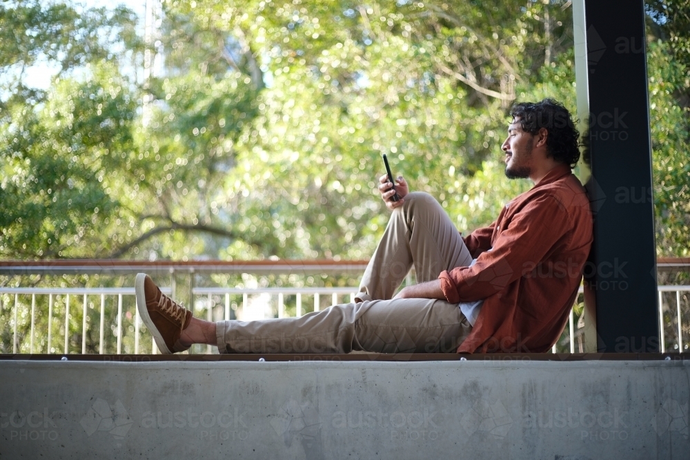 Image of Man sitting against pole on phone on an outdoor verandah ...