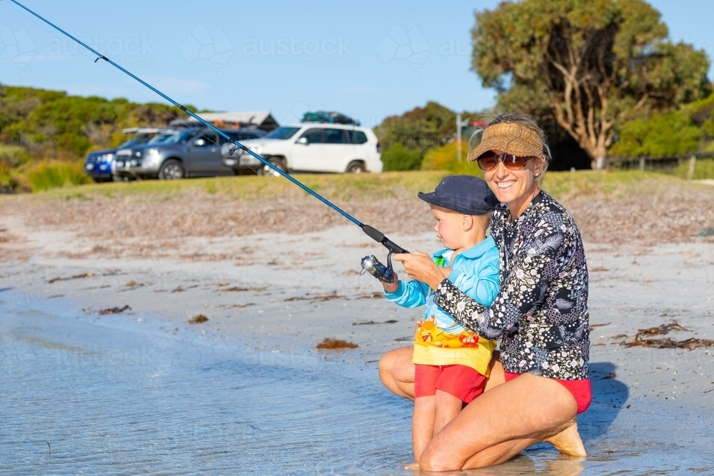Image of Mother helping toddler with fishing rod on beach - Austockphoto