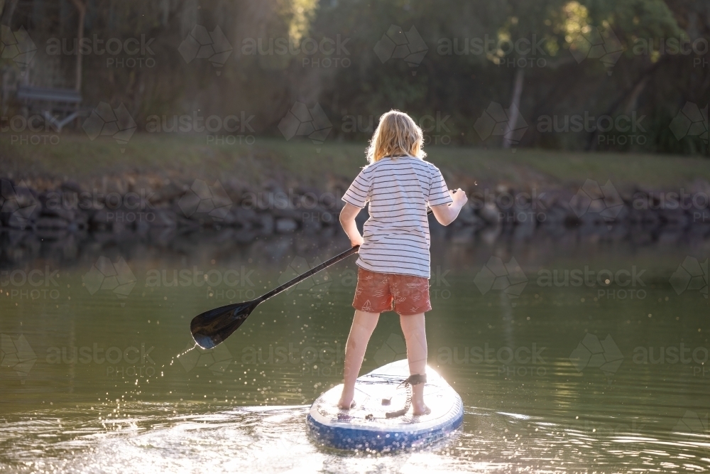 Image of Pre teen boy stand up paddle boarding on lake in golden ...
