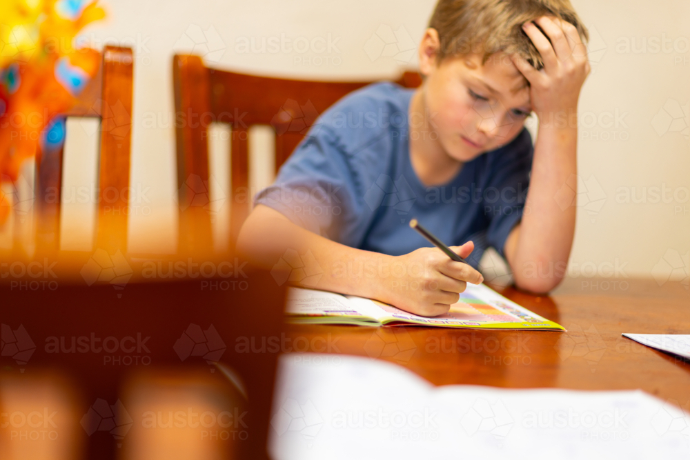 Image of school-aged boy with pencil in hand working on a problem in a workbook sitting at table ...