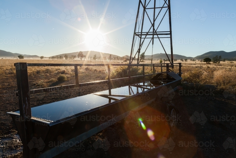 Image of Sun flare over water trough on farm with windmill - Austockphoto