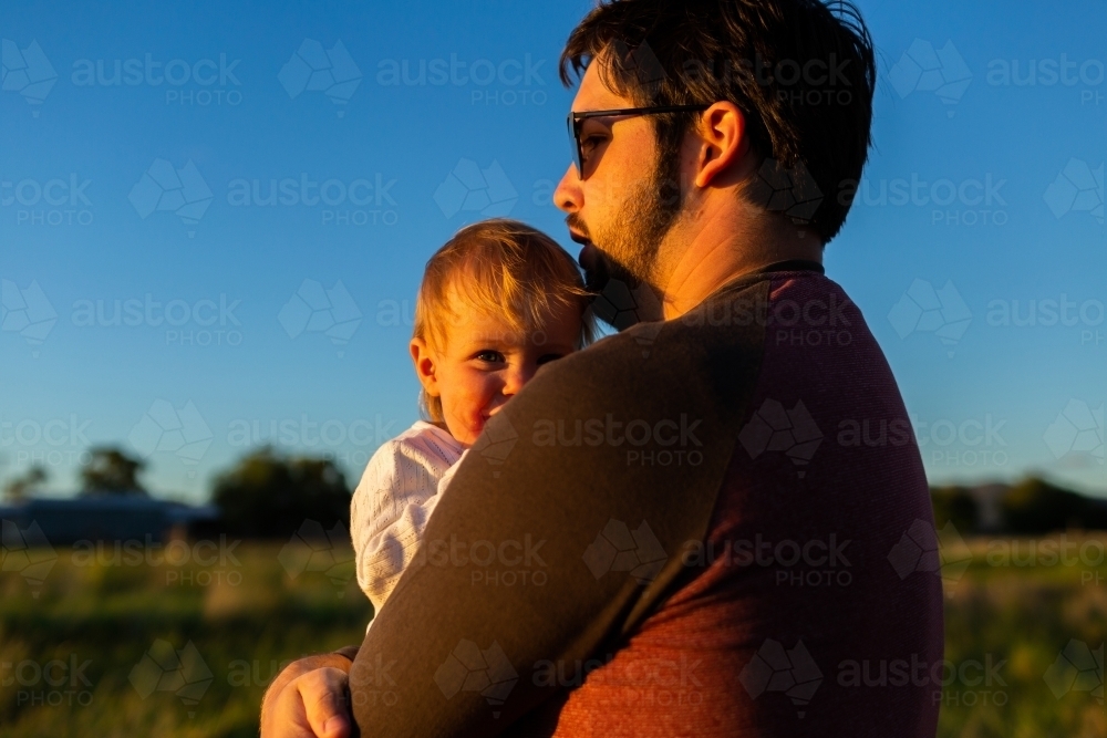Image of Sunlit man on farm hugging baby girl in arms standing in ...