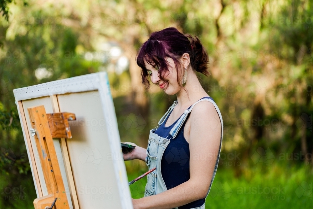 Image of Teen artist with purple hair working on painting in overalls ...