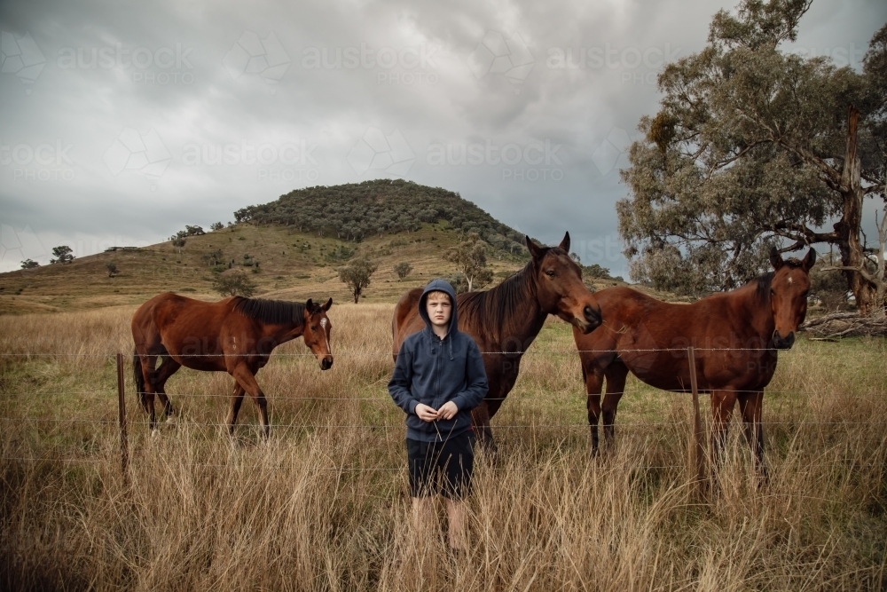 Image of Teen boy standing at farm fence with horses behind him ...