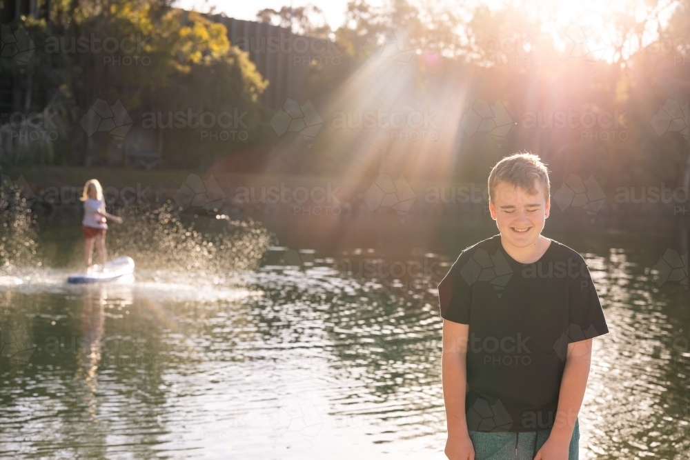 Image of Teenage boy standing by lake with younger brother on paddle ...