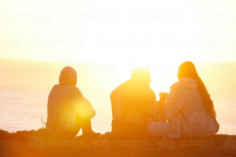 Image of Three people sitting by the beach at sunset. - Austockphoto