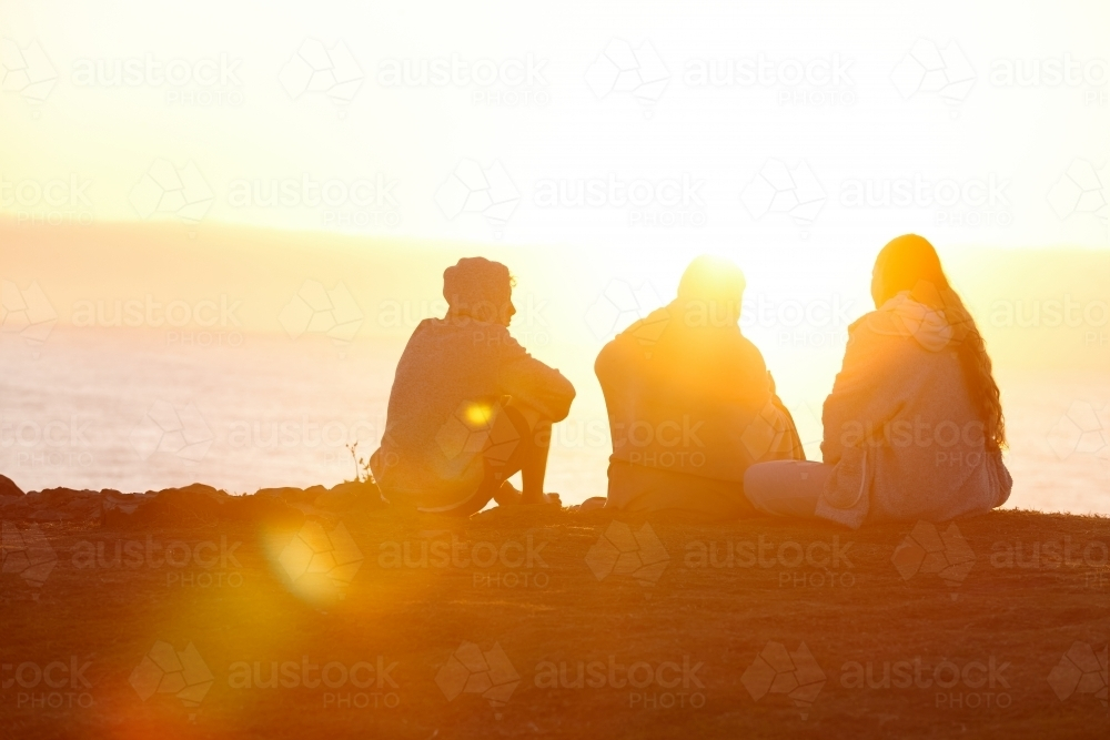 Image of Three people sitting by the beach at sunset - Austockphoto