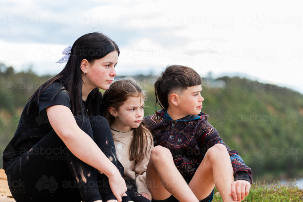 Image of Three siblings looking away together sitting on clifftop ...