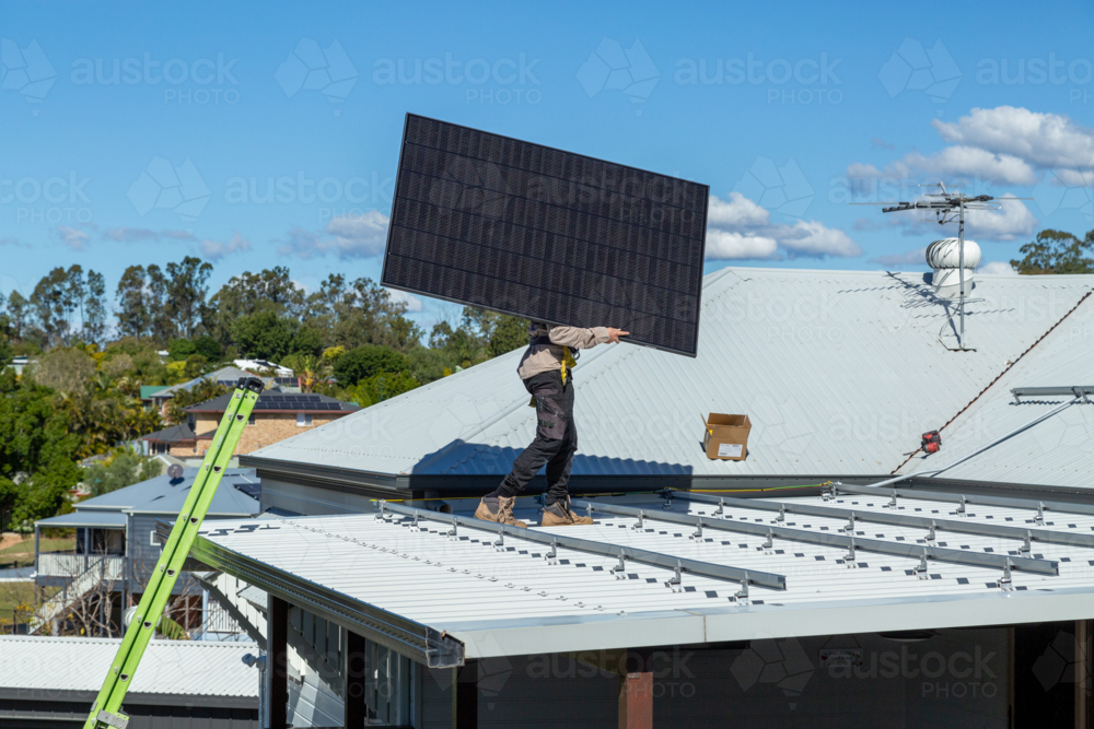 Image of Tradesman carrying solar panel onto metal roof of suburban ...