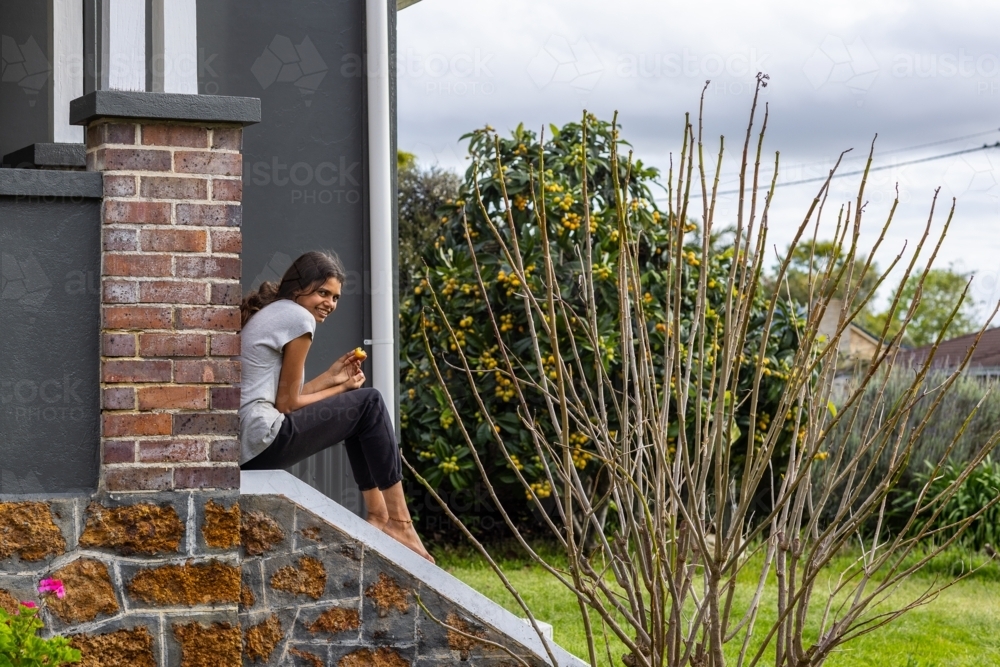 Image of tween girl sitting on steps of home eating a loquat from the ...