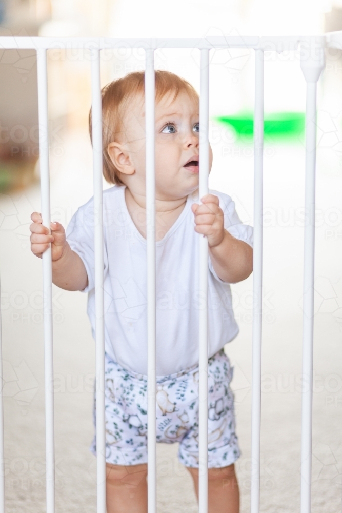 Image of Unsure looking child standing at baby gate fence holding bars ...