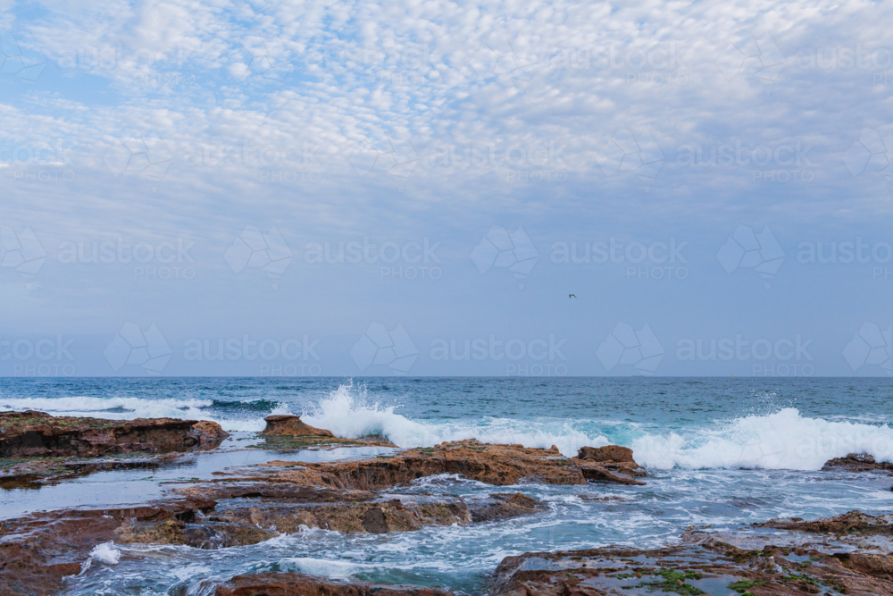 Image of Waves splash against coastal rocks under soft blue clouded sky ...