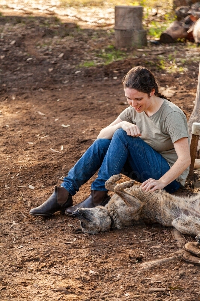 Image of Woman sitting on ground under gum tree scratching dog ...