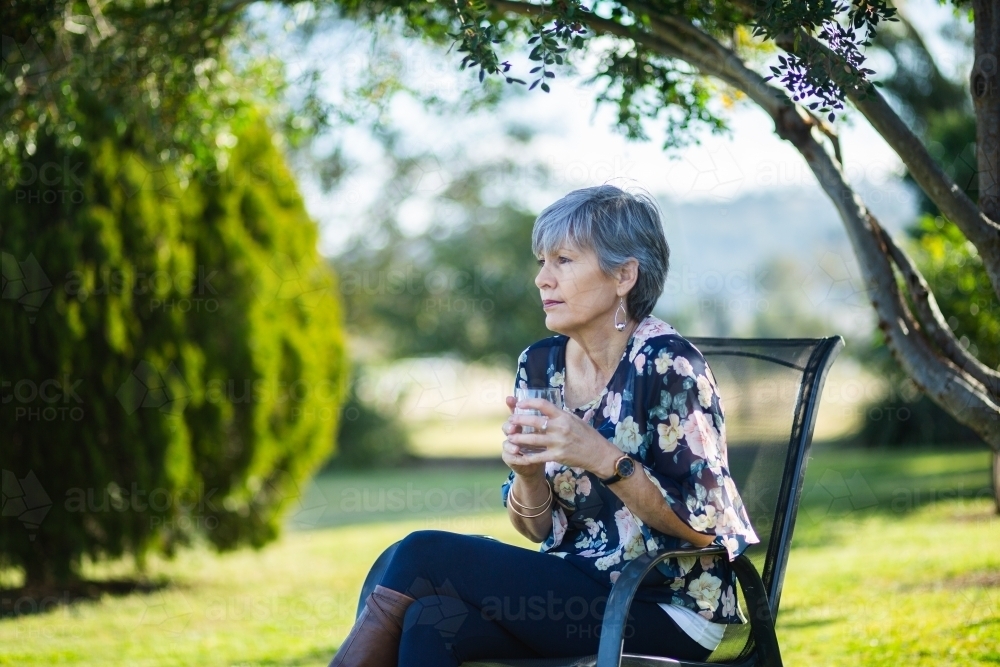 Image of Woman with a glass of water sitting in chair under shady tree ...