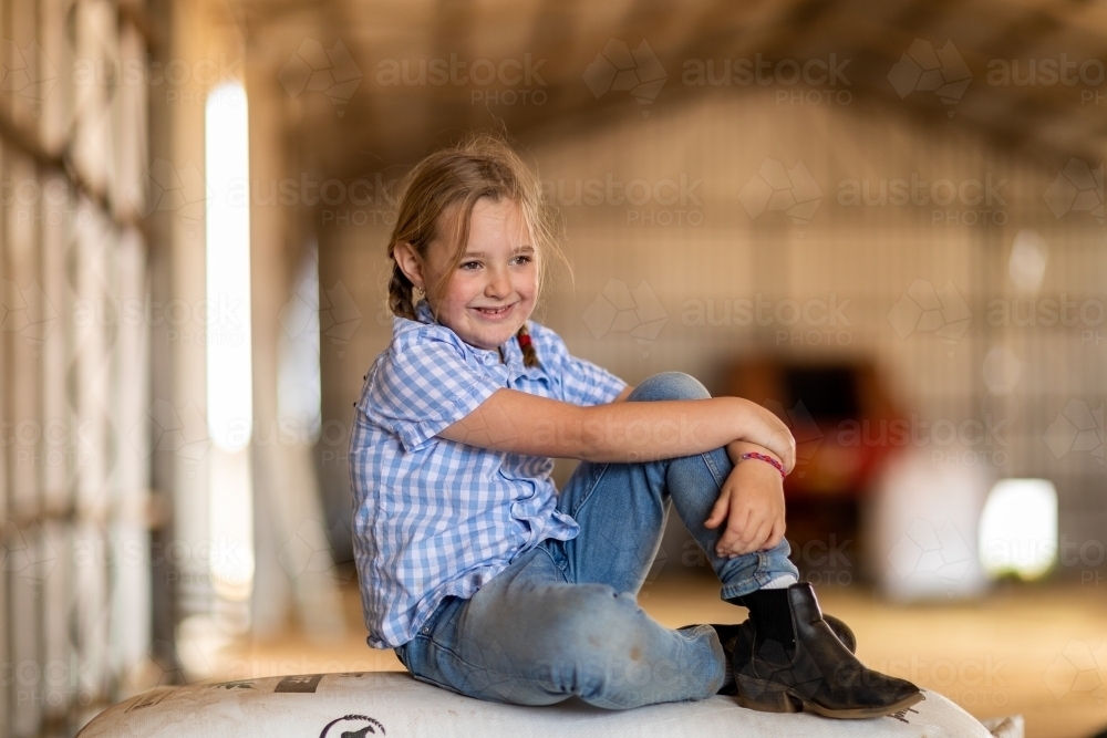 Image of young farm girl sitting on chaff bale in farm shed - Austockphoto