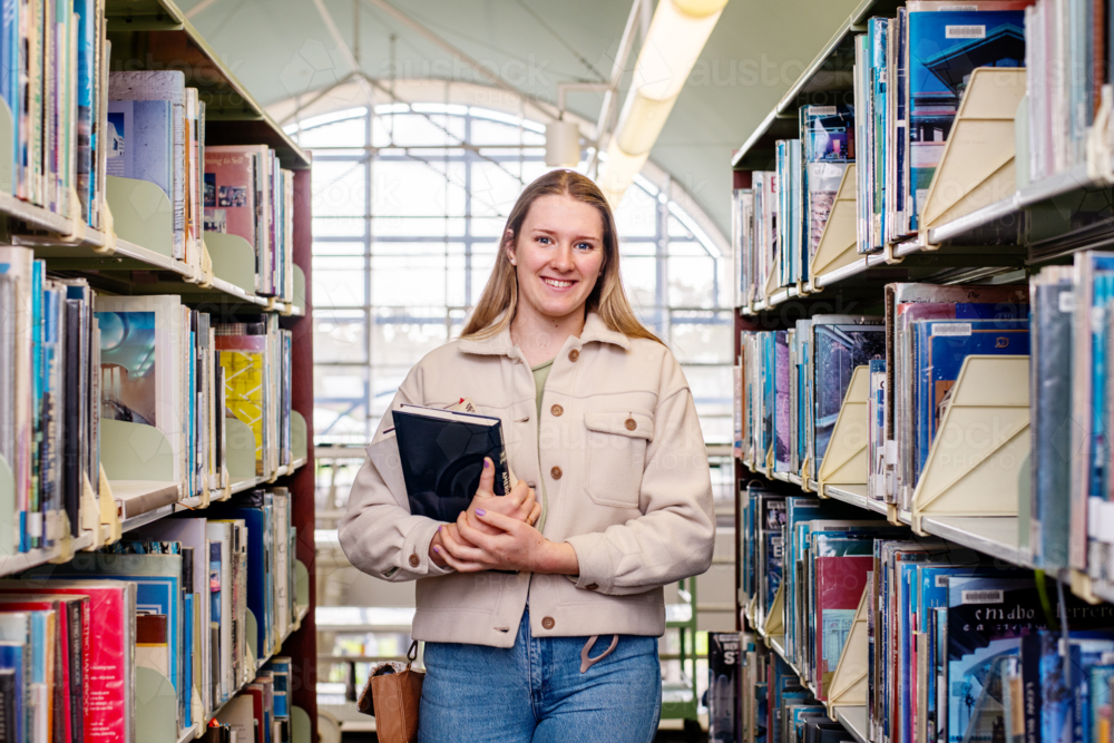 Image of Young woman holding book in uni library with bookshelves ...